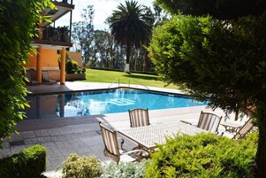 Uma área de piscina com espreguiçadeiras e uma mesa, cercada por vegetação e um edifício com varanda.