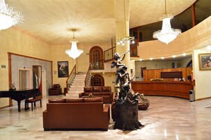 Um hall amplo com um piano de cauda, sofás de couro, um balcão de recepção e um lustre.
