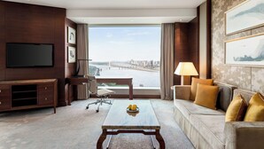 Quarto de hotel moderno com uma grande janela oferecendo vista da cidade, televisão de tela plana, mesa de jantar com cesta de frutas e sofá com almofadas.