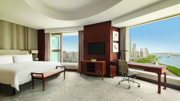 Quarto de hotel com uma cama grande, uma mesa, uma cadeira, uma televisão e vista para a paisagem urbana e para a água.