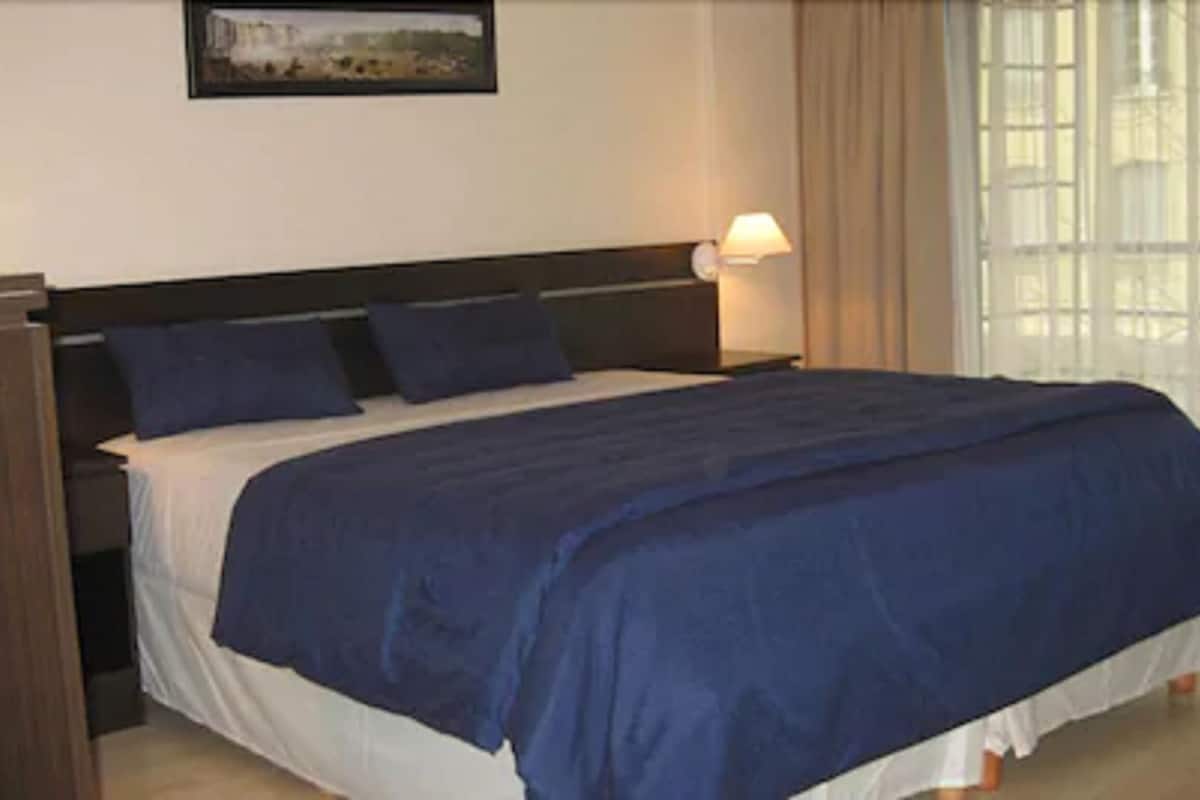 Quarto de hotel com uma cama grande, uma mesa de cabeceira, um abajur e uma janela com cortinas.