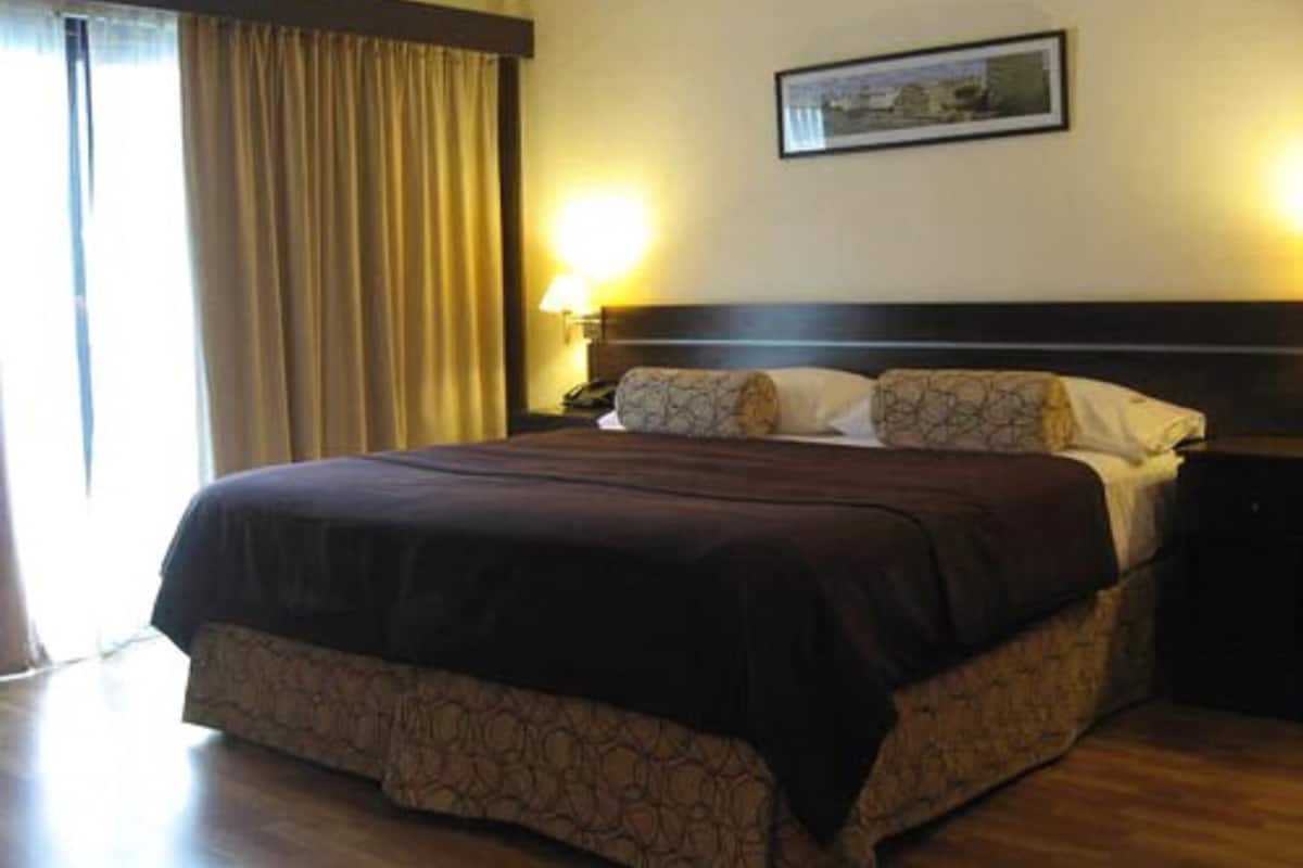 Quarto de hotel com uma cama grande, mesa de cabeceira, abajur e um quadro na parede.