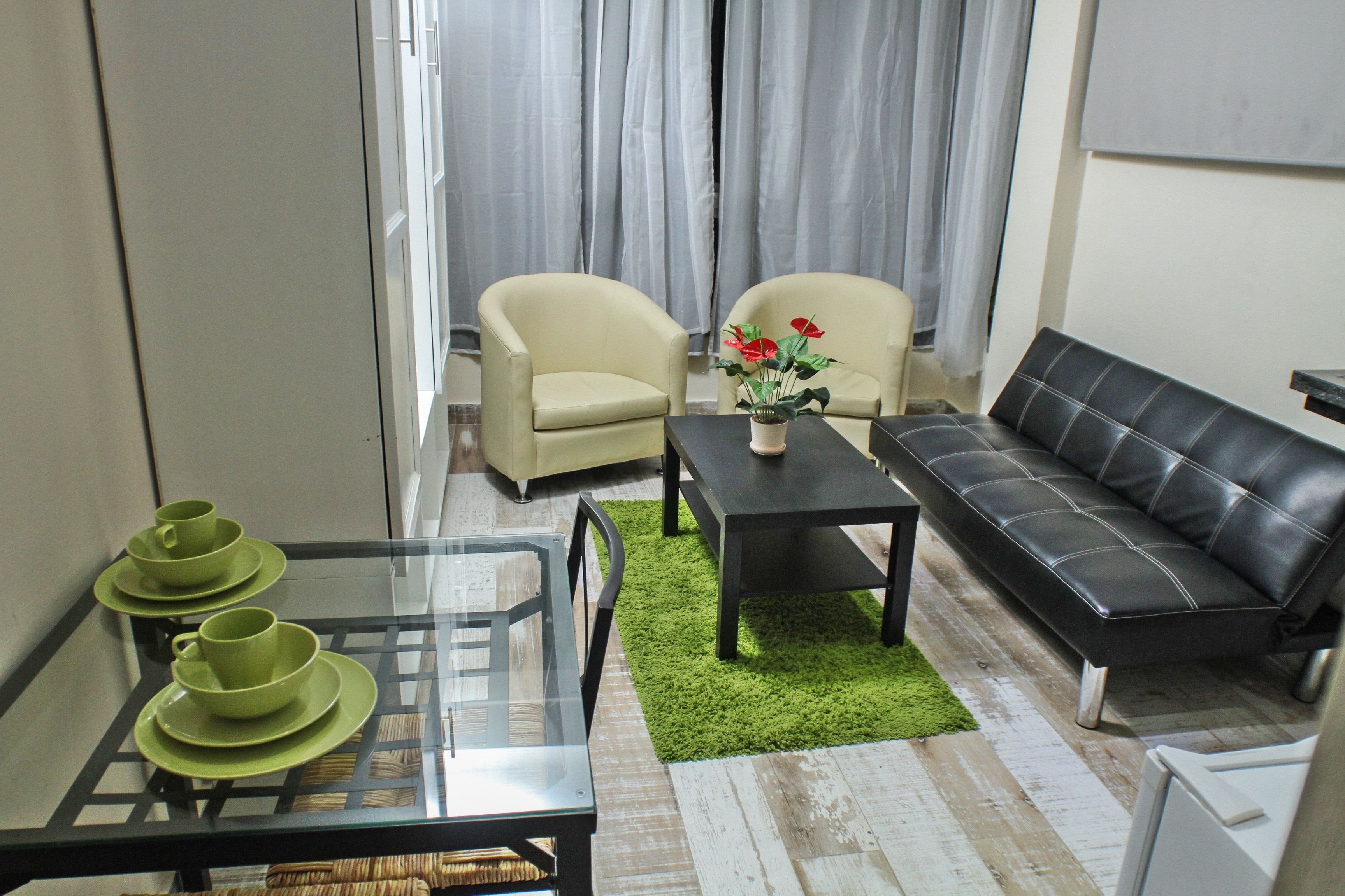 Pequena sala de estar com mesa de vidro, duas cadeiras, sofá de couro preto e tapete verde.