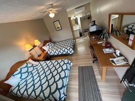 Quarto de hotel com duas camas, uma escrivaninha, uma cadeira, um espelho, um ventilador de teto e um banheiro visível através de uma porta aberta.