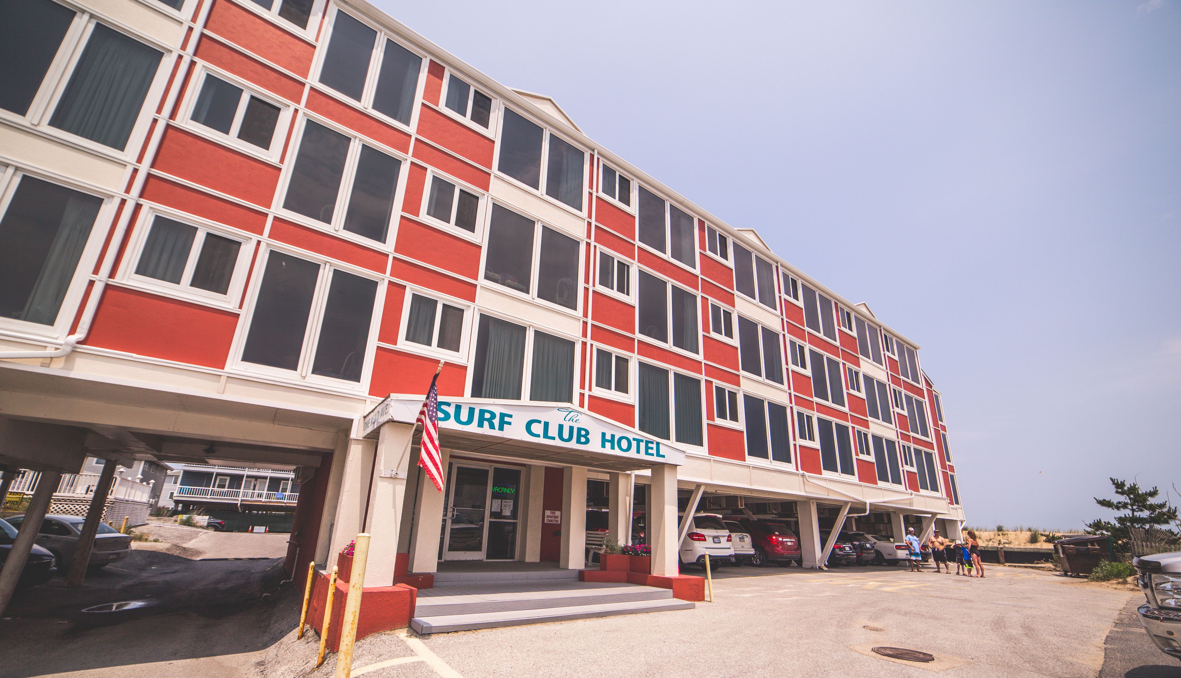 Um hotel com vários andares, fachada em vermelho e branco, um grande letreiro 'Surf Club Hotel' e um estacionamento com vários carros.