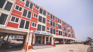 Um hotel com vários andares, fachada em vermelho e branco, um grande letreiro 'Surf Club Hotel' e um estacionamento com vários carros.