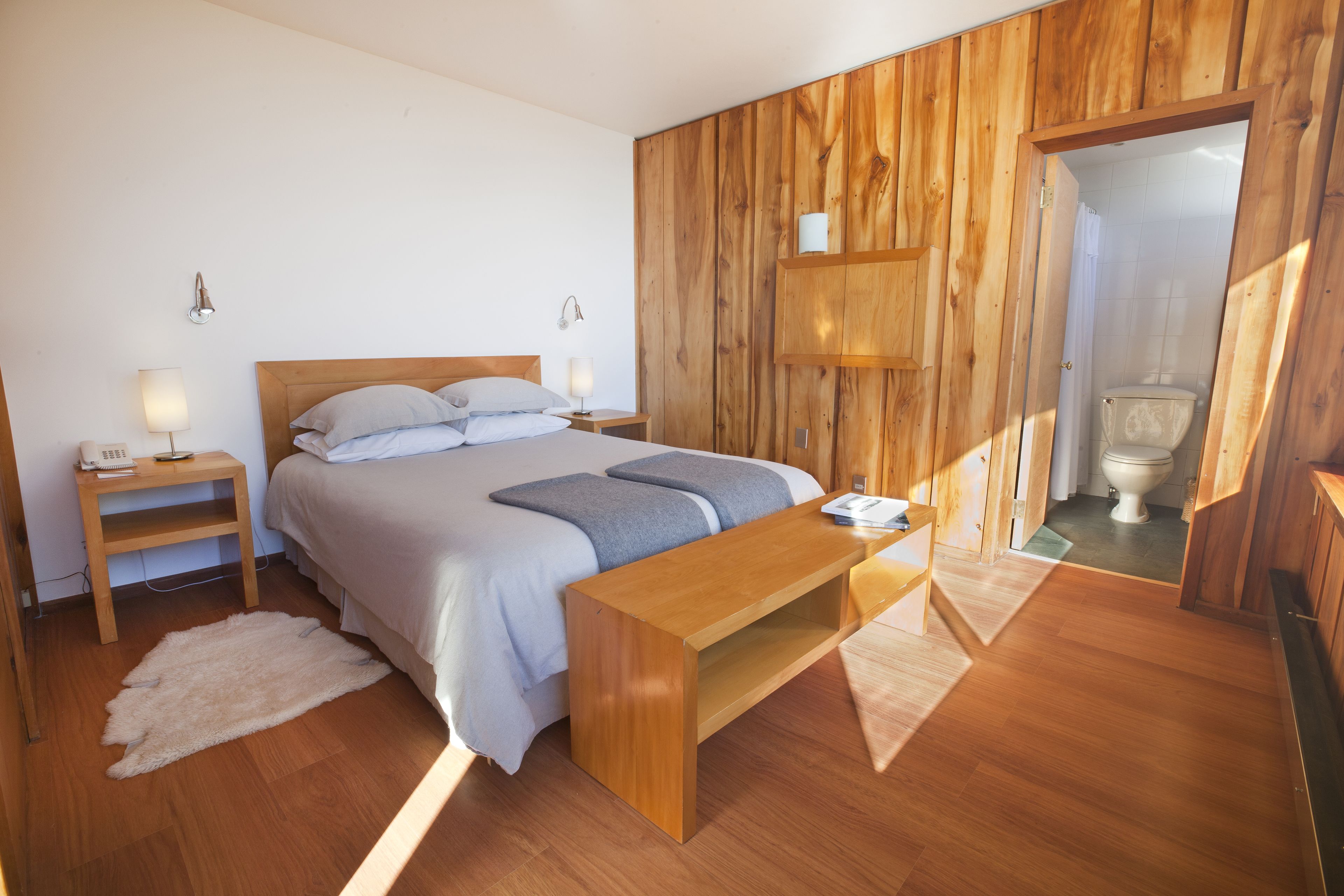 Quarto com painel de parede de madeira, cama com almofadas, mesa de cabeceira com abajur e um banco de madeira.