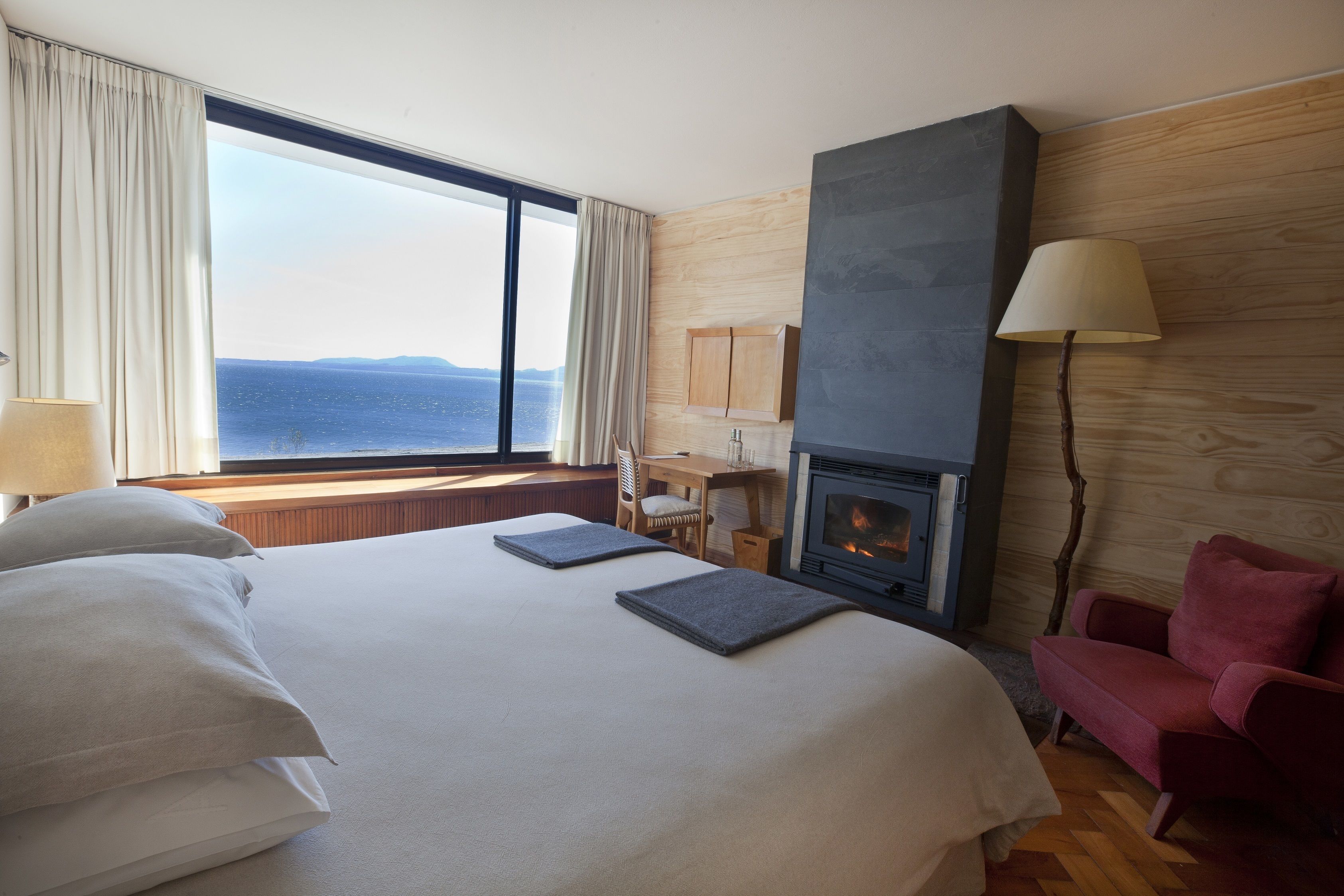 Quarto de hotel com uma cama grande, uma escrivaninha, uma cadeira, uma poltrona vermelha, uma lareira e vista para o mar.