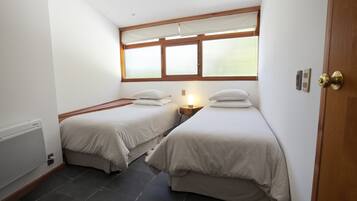 Quarto com duas camas, janela com persianas, radiador e porta de madeira.