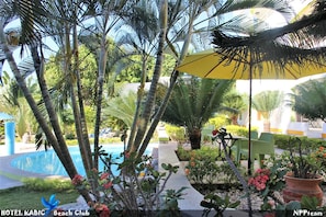 Área de piscina com palmeiras, um guarda-sol amarelo e um caminho que leva a um edifício.