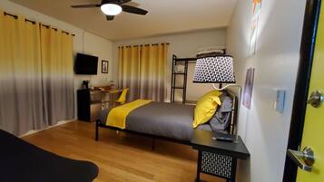 Quarto com cama, escrivaninha, cadeira, ventilador de teto e televisão.