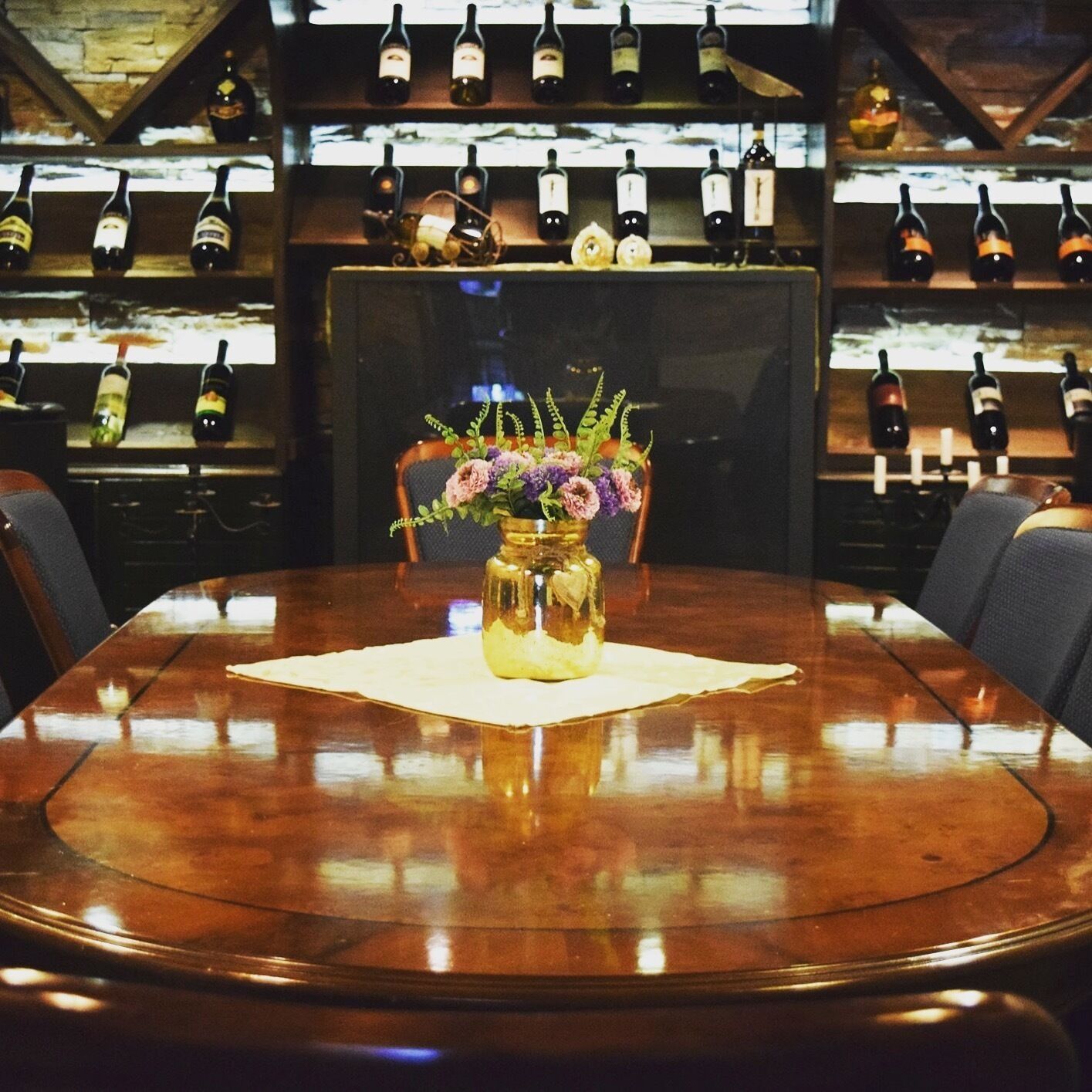 Uma mesa redonda de madeira com um vaso de vidro cheio de flores no centro, rodeada por cadeiras. Atrás da mesa, há uma parede com prateleiras que exibem diversas garrafas, provavelmente de vinho.