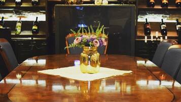 Uma mesa redonda de madeira com um vaso de vidro cheio de flores no centro, rodeada por cadeiras. Atrás da mesa, há uma parede com prateleiras que exibem diversas garrafas, provavelmente de vinho.