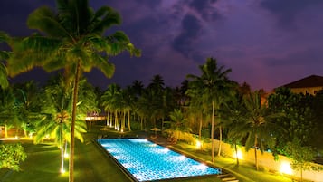 Vista noturna de uma piscina rodeada por palmeiras e caminhos bem iluminados.