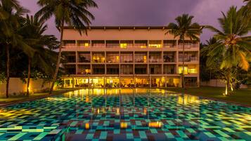 Um hotel com piscina apresentando padrão quadriculado, rodeado por palmeiras e um edifício de vários andares com sacadas.