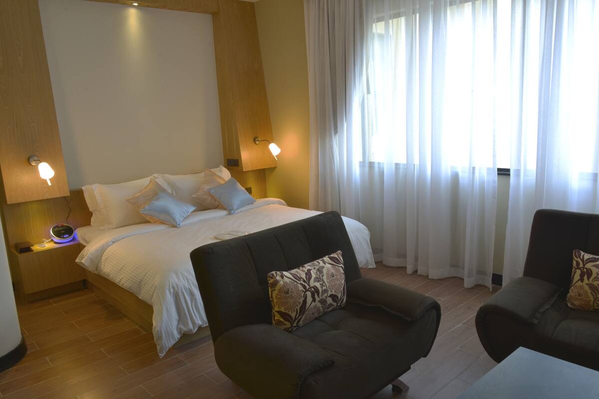 Quarto de hotel com cama, duas luminárias de cabeceira, um sofá e uma janela com cortinas.