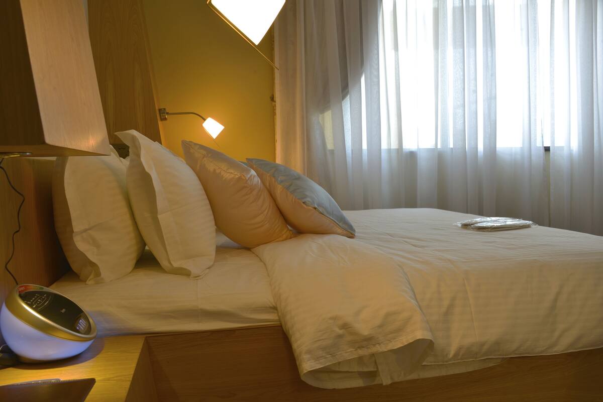 Uma cama bem arrumada com roupa de cama branca, uma mesa de cabeceira com abajur e cortinas leves na janela.