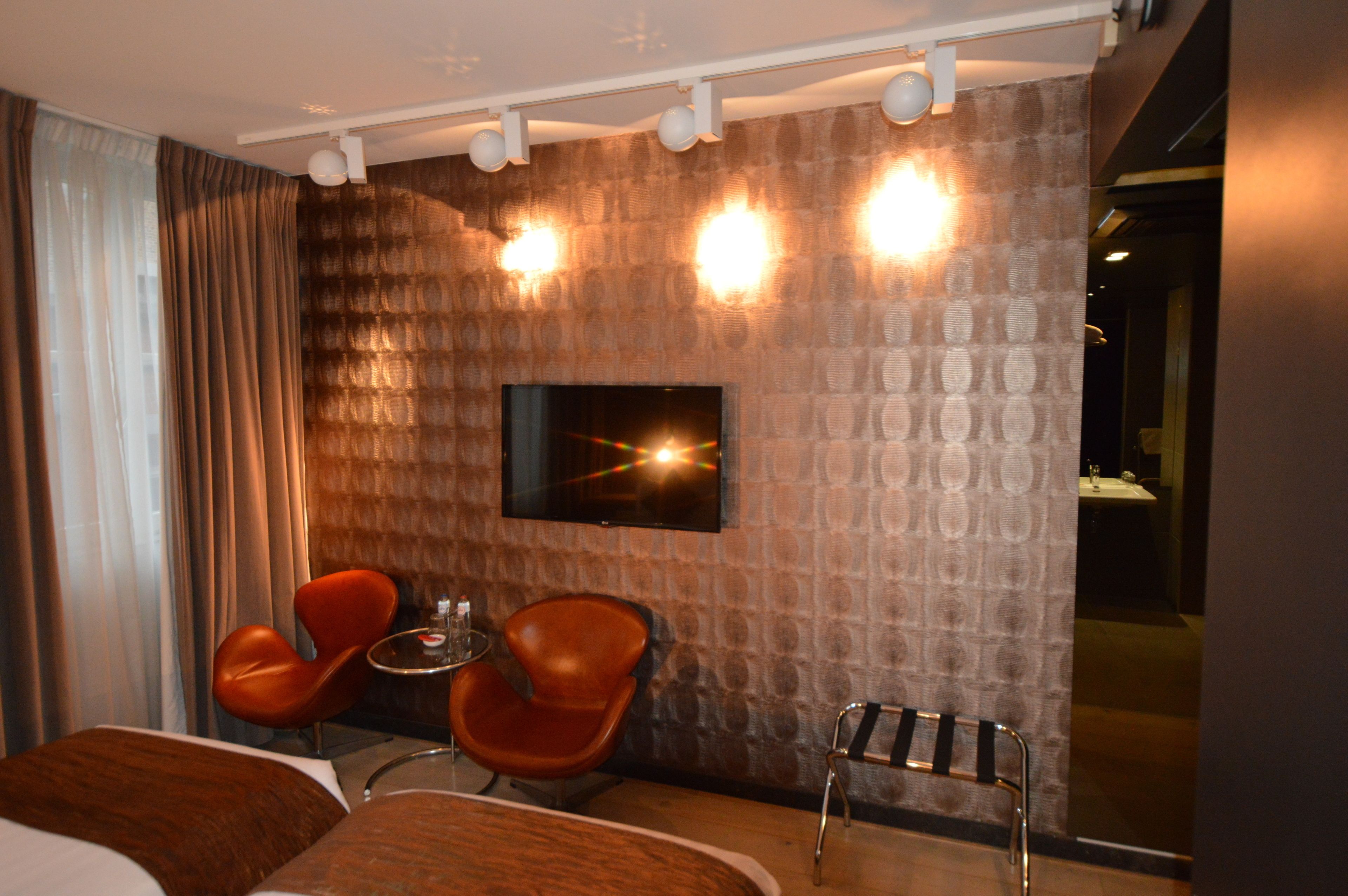 Quarto de hotel com uma televisão de tela plana grande fixada em uma parede texturizada, duas poltronas de couro, uma mesa redonda pequena e uma cama com cabeceira de madeira.
