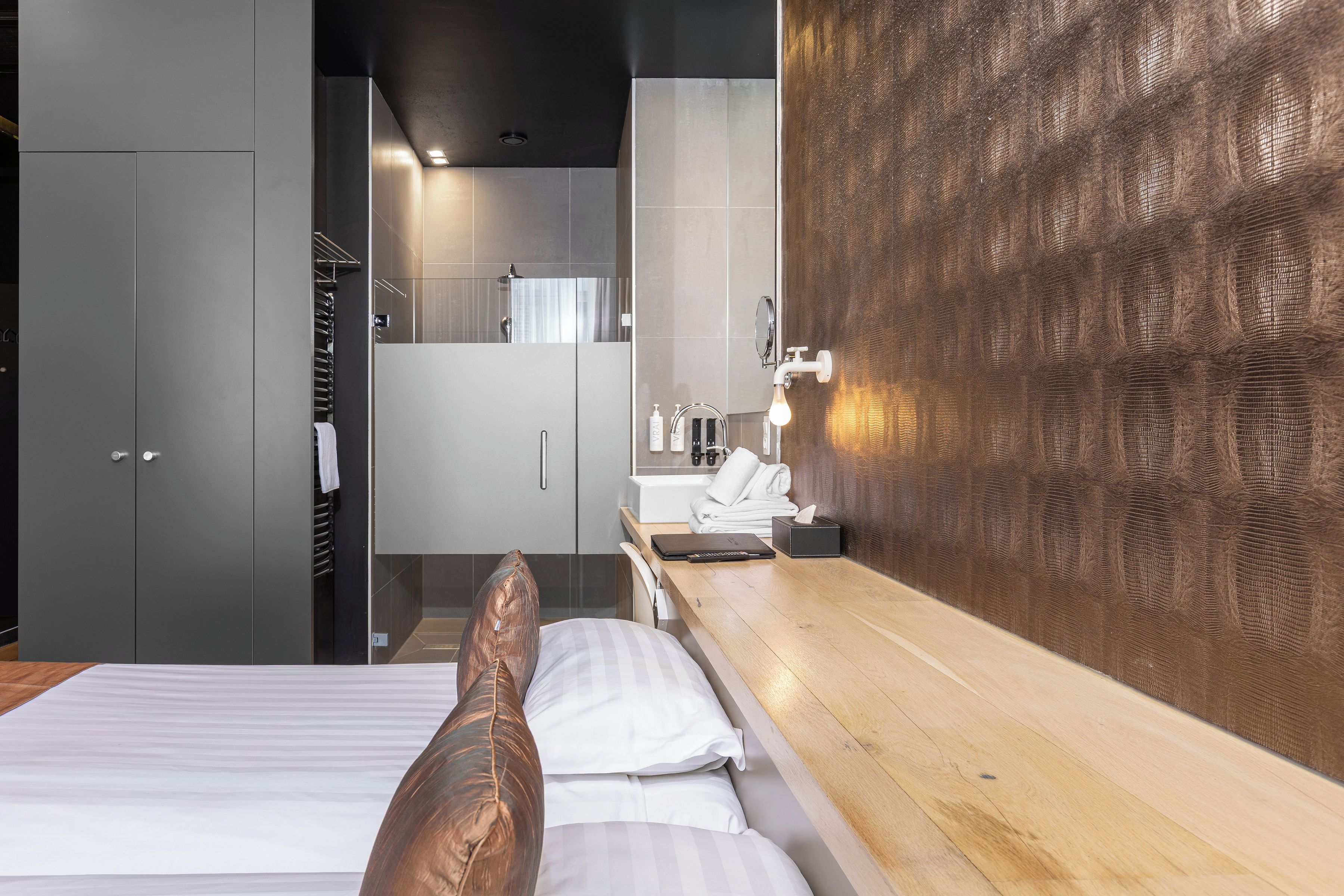 Quarto de hotel moderno com uma cama, mesa de cabeceira de madeira, espelho e banheiro com chuveiro e porta-toalhas.