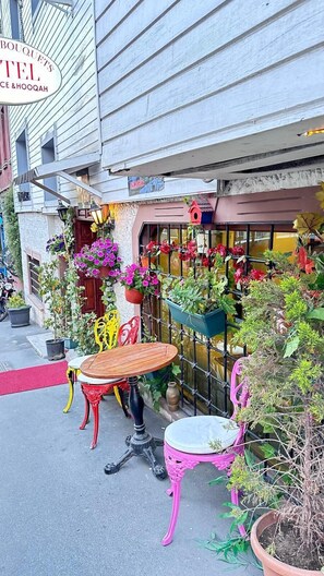 Exterior de hotel com toldo vermelho, mesas e cadeiras ao ar livre, e cestos de flores pendurados.