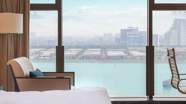 Quarto de hotel com uma cama grande, uma cadeira e vista para a paisagem urbana e para a água.