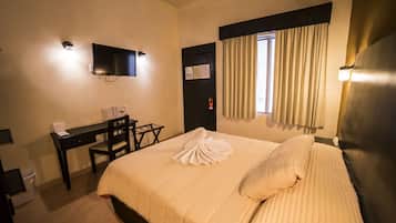 Quarto de hotel com cama, escrivaninha, cadeira, televisão e janela com cortinas.