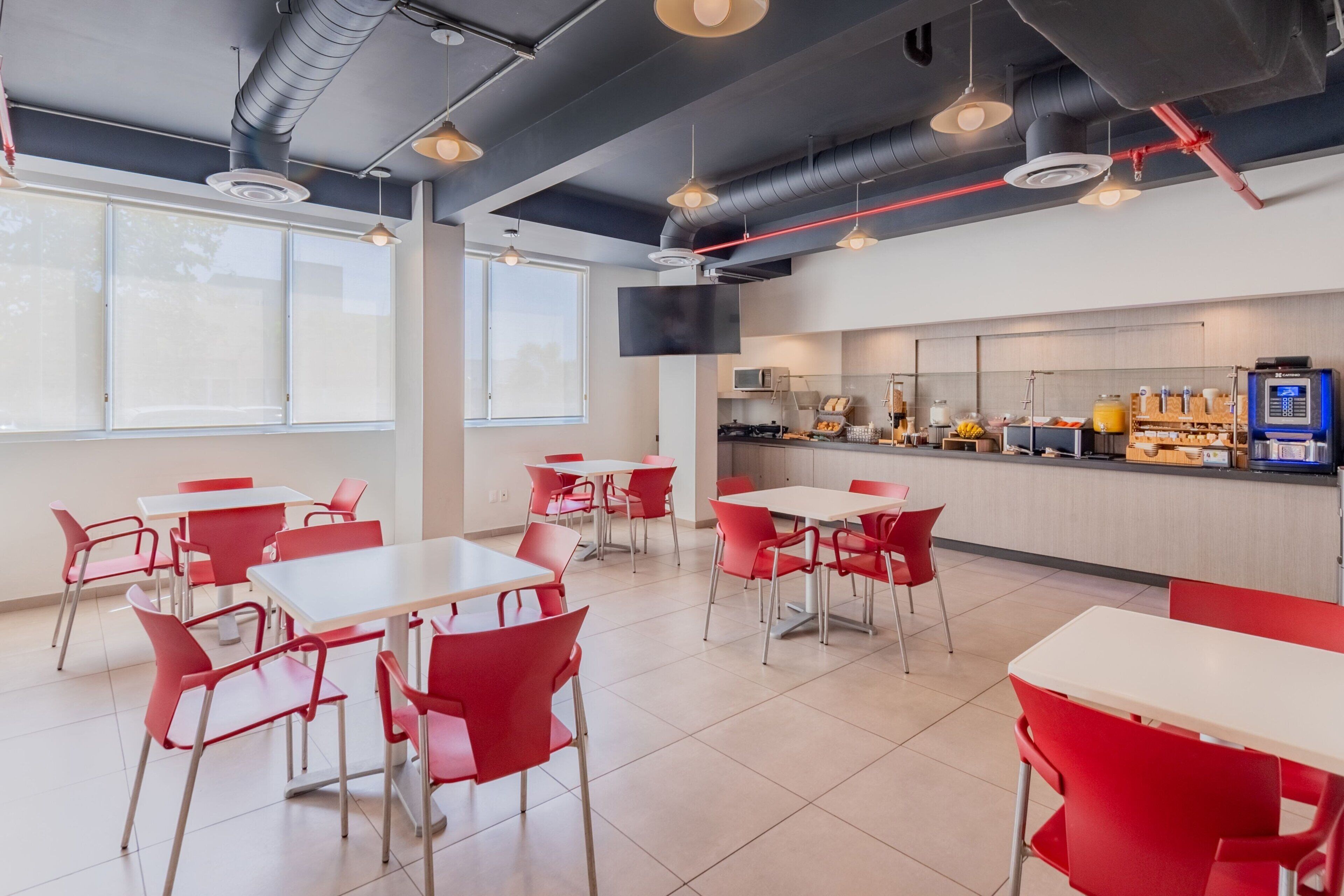 Uma sala de jantar moderna com cadeiras vermelhas e mesas brancas, janelas amplas e uma bancada de cozinha com diversos eletrodomésticos.