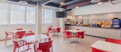 Uma sala de jantar moderna com cadeiras vermelhas e mesas brancas, janelas amplas e uma bancada de cozinha com diversos eletrodomésticos.
