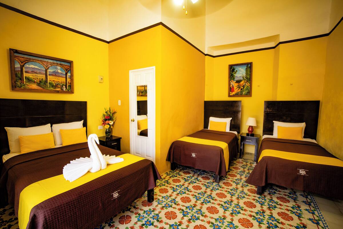 Quarto de hotel com quatro camas, cada uma com colcha marrom e almofadas brancas. O quarto tem paredes amarelas e piso estampado. Há quadros nas paredes e uma porta com espelho.