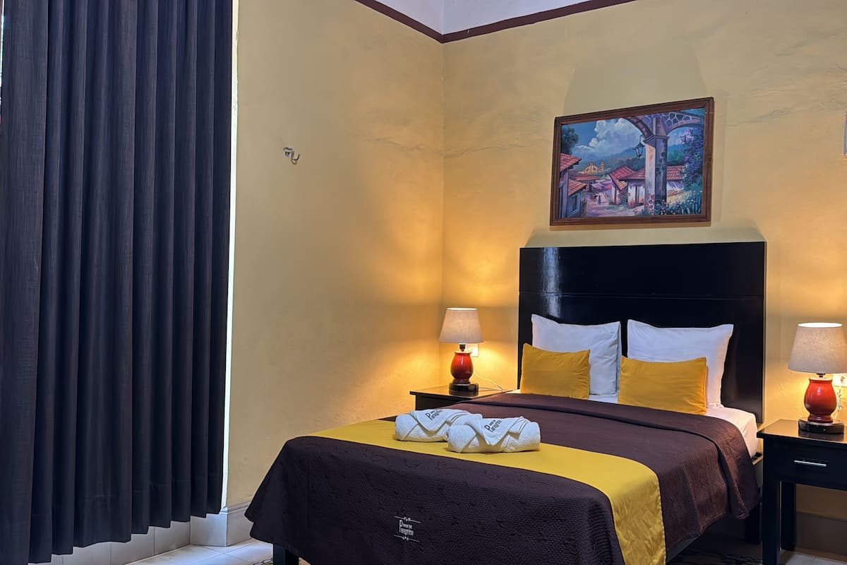 Quarto de hotel com cama, duas luminárias de cabeceira, um quadro na parede e uma janela com cortinas.