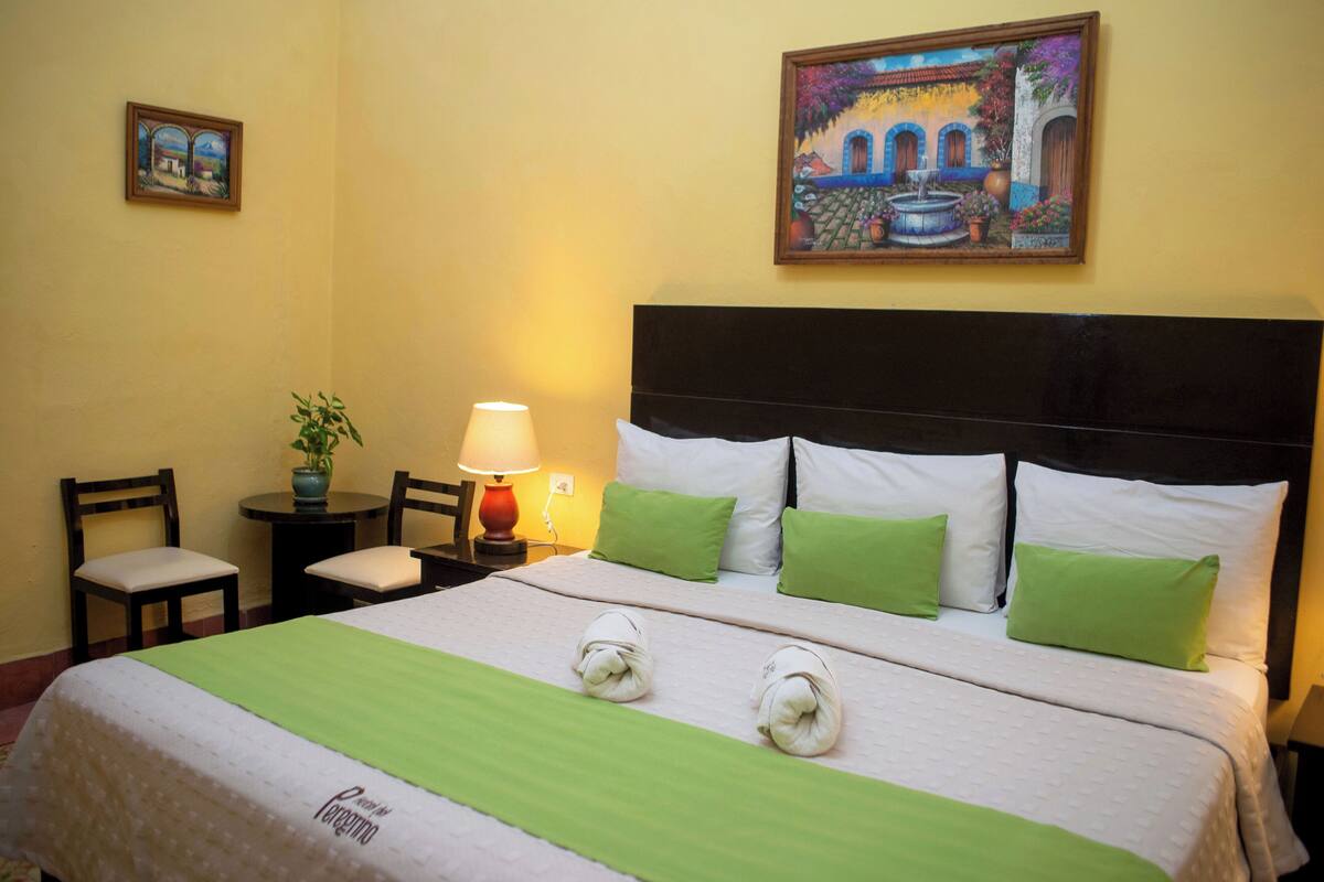 Quarto de hotel com uma cama, almofadas verdes, uma mesa de cabeceira com abajur, uma cadeira e um quadro na parede.