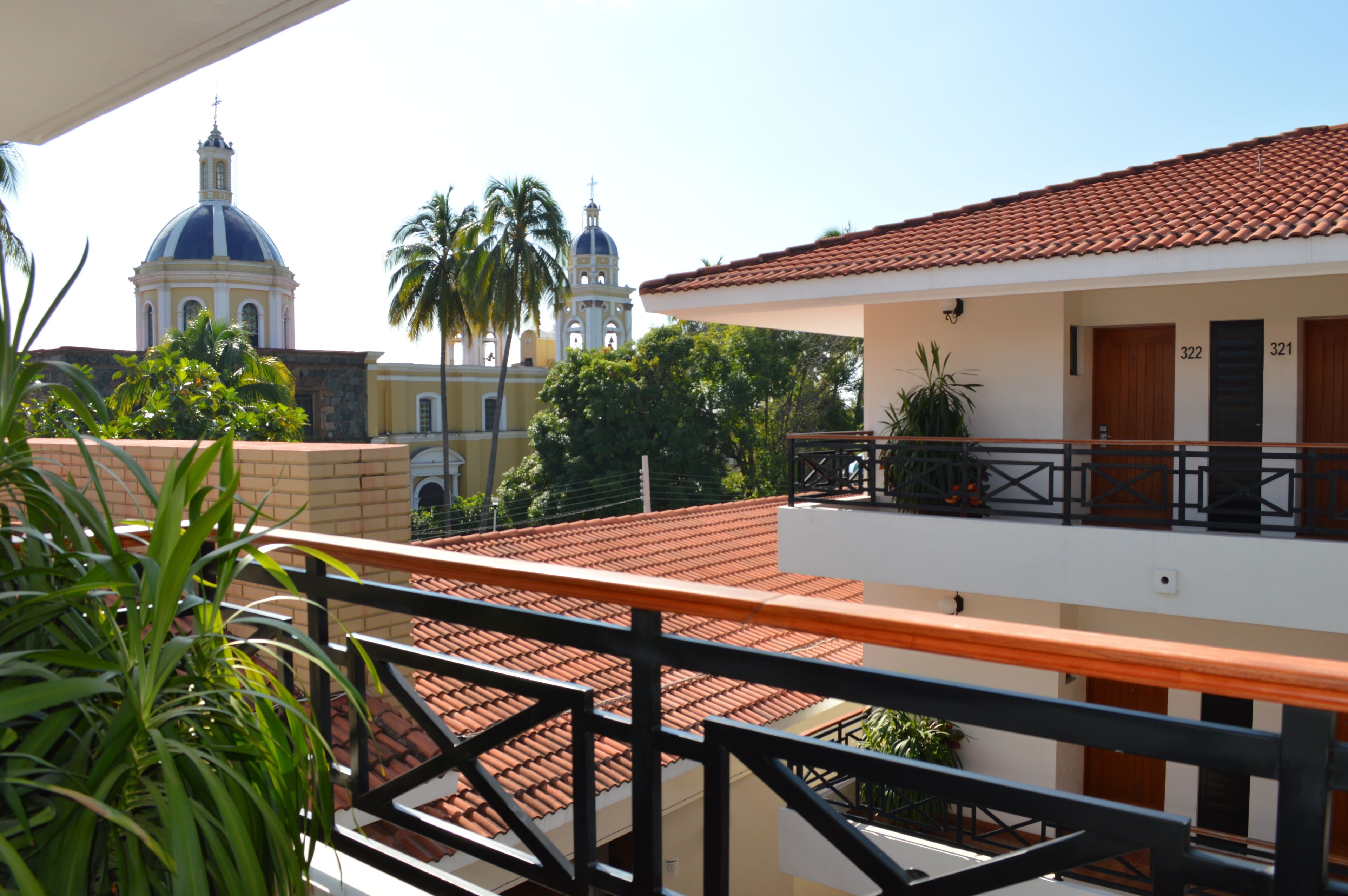 Um hotel com telhado de telhas vermelhas, varanda com guarda-corpo preto e vista para uma igreja com cúpula e campanário.
