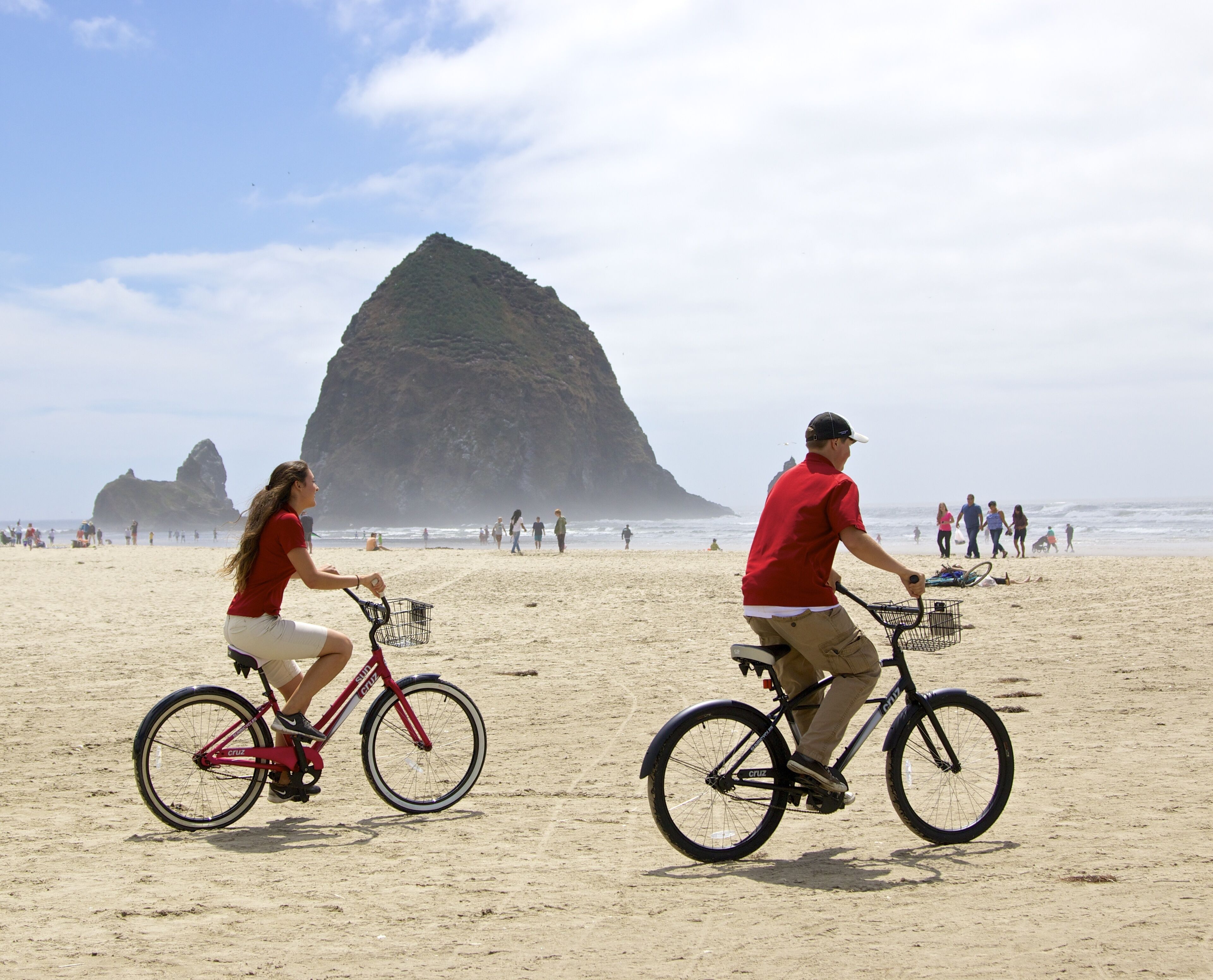 Duas pessoas andando de bicicleta em uma praia arenosa, com uma grande formação rochosa ao fundo.