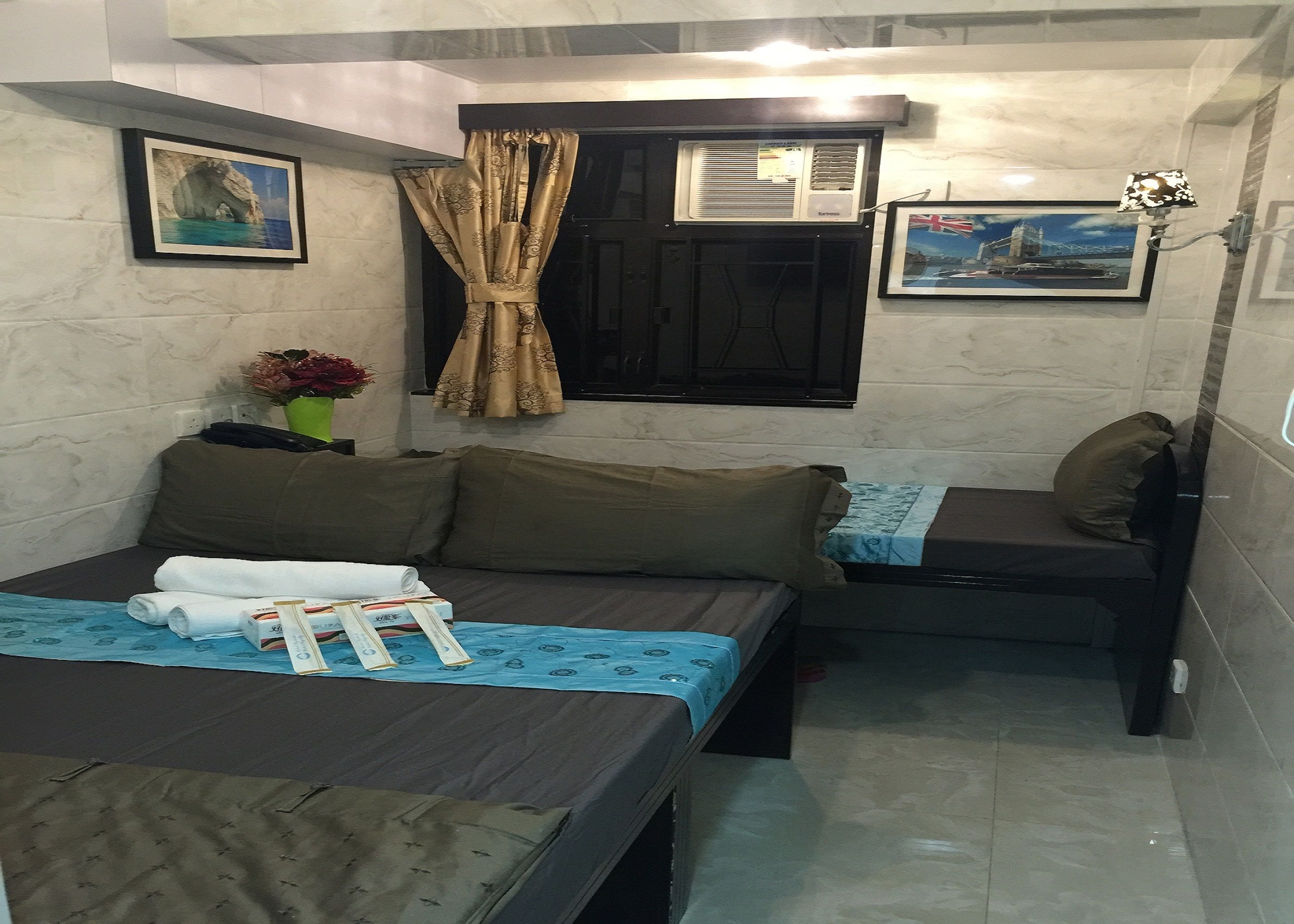 Um quarto pequeno e acolhedor, com uma cama, um banco e uma janela com cortinas. Há quadros nas paredes e um abajur sobre o banco.