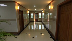 Corredor de hotel com pisos lustrosos, portas de madeira e luminárias fixadas na parede.