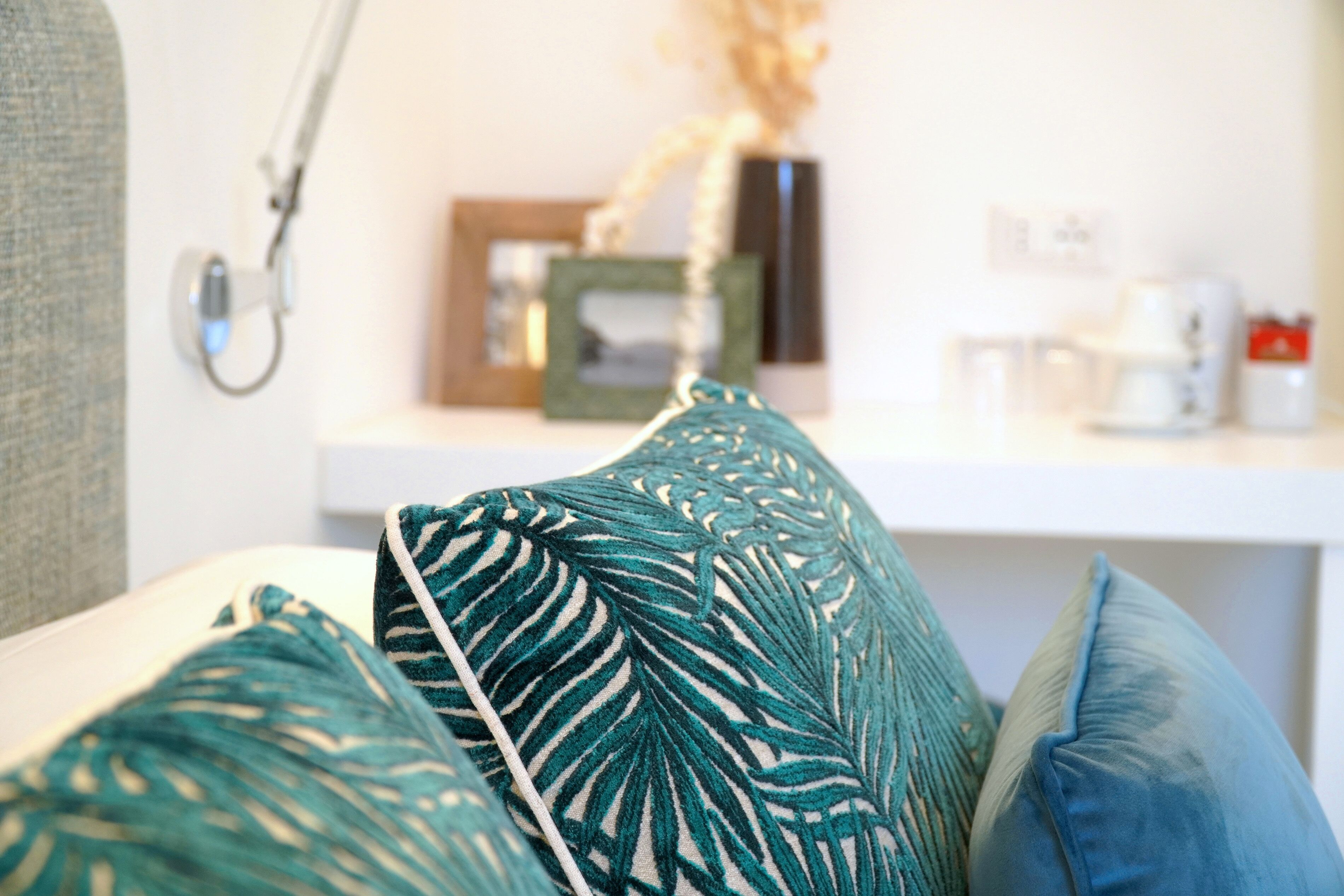 Uma cama com almofadas estampadas em tons de verde-água e azul, uma mesa de cabeceira branca com um quadro emoldurado, um vaso com plantas secas e uma cafeteira.