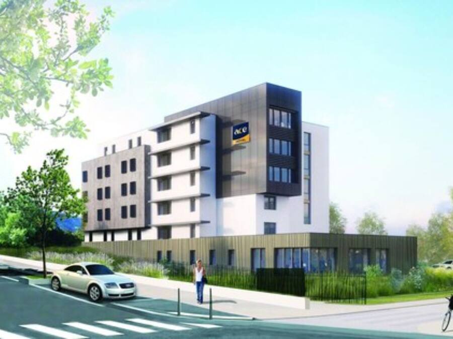 Um edifício moderno de vários andares com telhado plano e um destaque para a placa 'ACE' na lateral, com uma faixa de pedestres na frente.