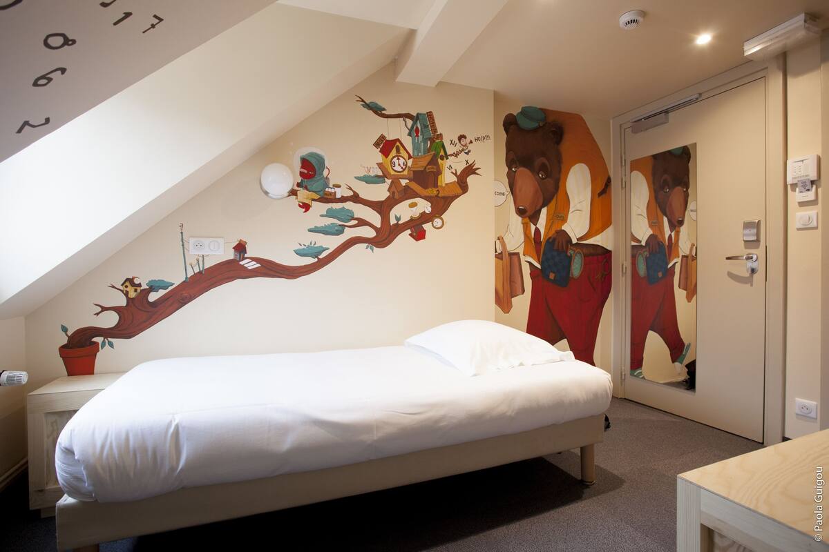 Quarto de hotel com cama, mesa de cabeceira, painel decorativo na parede e porta com espelho.
