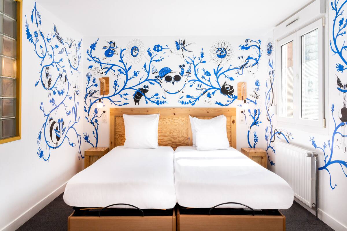Uma cama de casal com roupa de cama branca, uma cabeceira de madeira e uma parede com adesivos florais e de animais azuis.