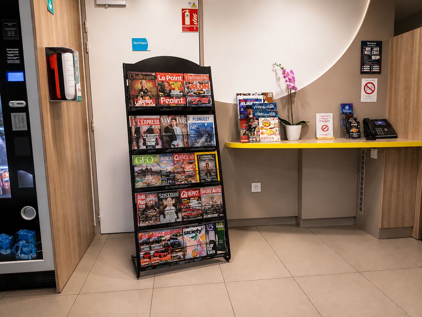 Uma estante com diversas publicações, uma máquina de venda automática e uma mesa de recepção com telefone e uma pequena planta em vaso.