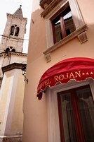 Um hotel chamado "Hotel Rojan", com toldo vermelho e uma torre de sino ao fundo.