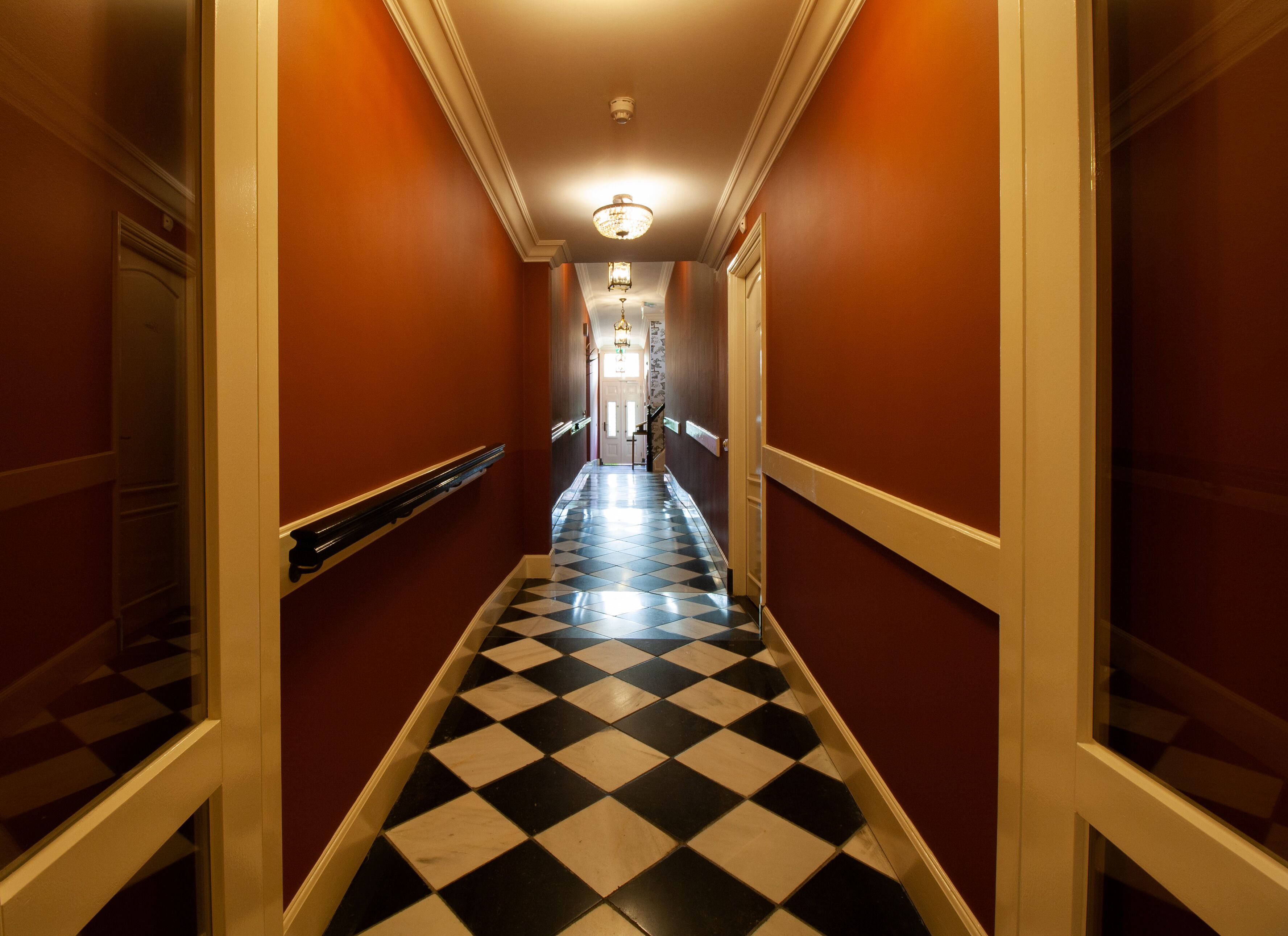 Corredor de hotel com chão quadriculado, paredes vermelhas e detalhes em branco.
