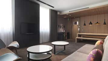 Quarto de hotel moderno com sofá, duas mesas redondas, televisão de tela plana e parede decorativa em madeira.