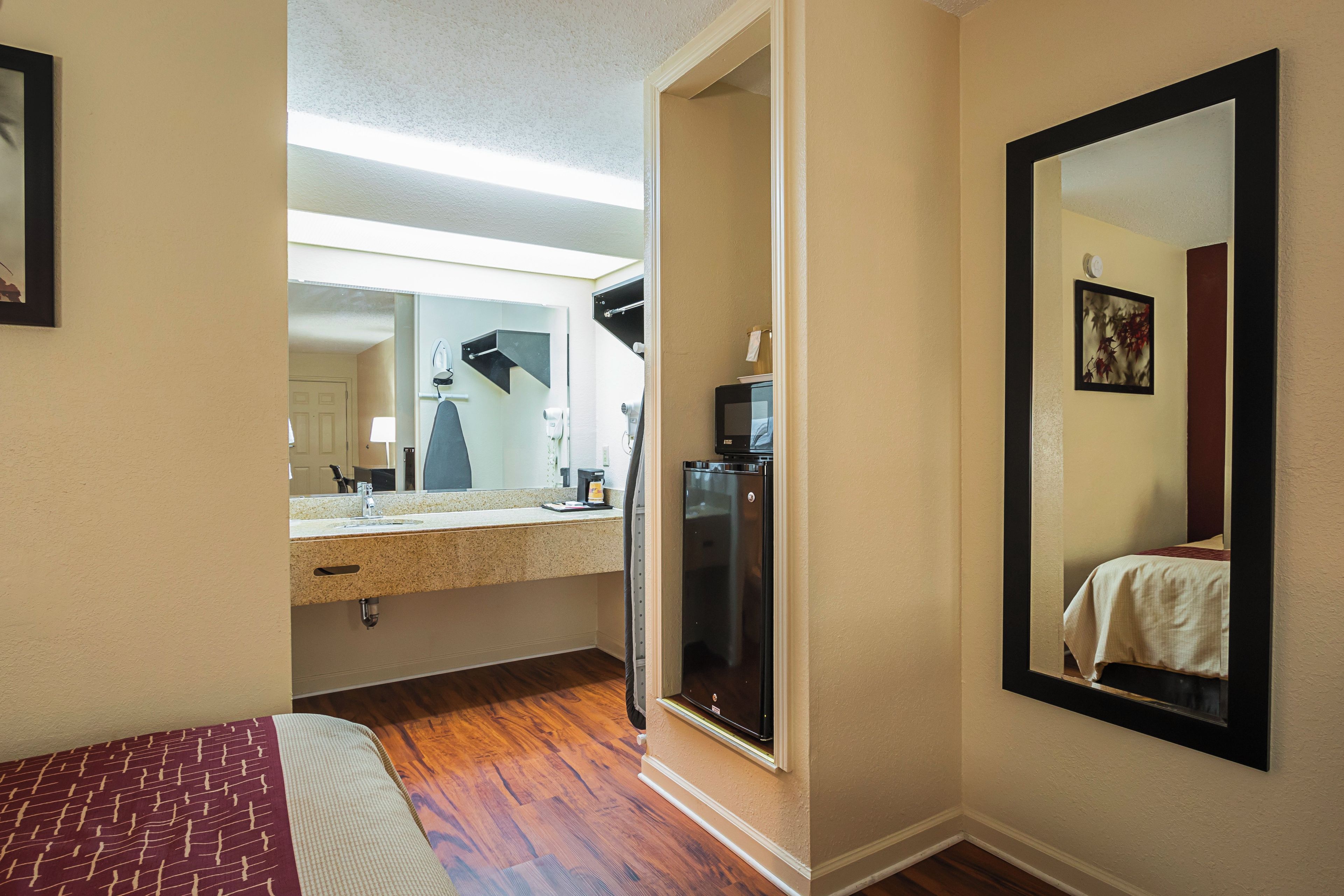 Quarto de hotel com cama, escrivaninha, espelho, geladeira e janela.