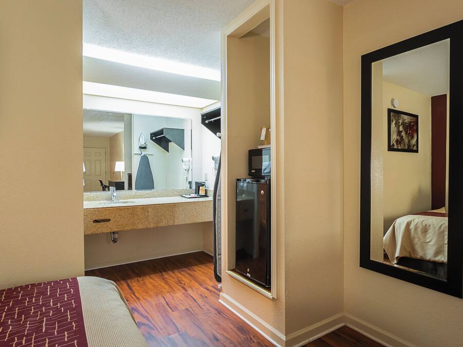 Quarto de hotel com cama, escrivaninha, espelho, geladeira e janela.