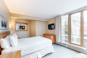 Quarto de hotel com uma cama grande, mesa de cabeceira de madeira, televisão na parede e uma janela ampla com cortinas.