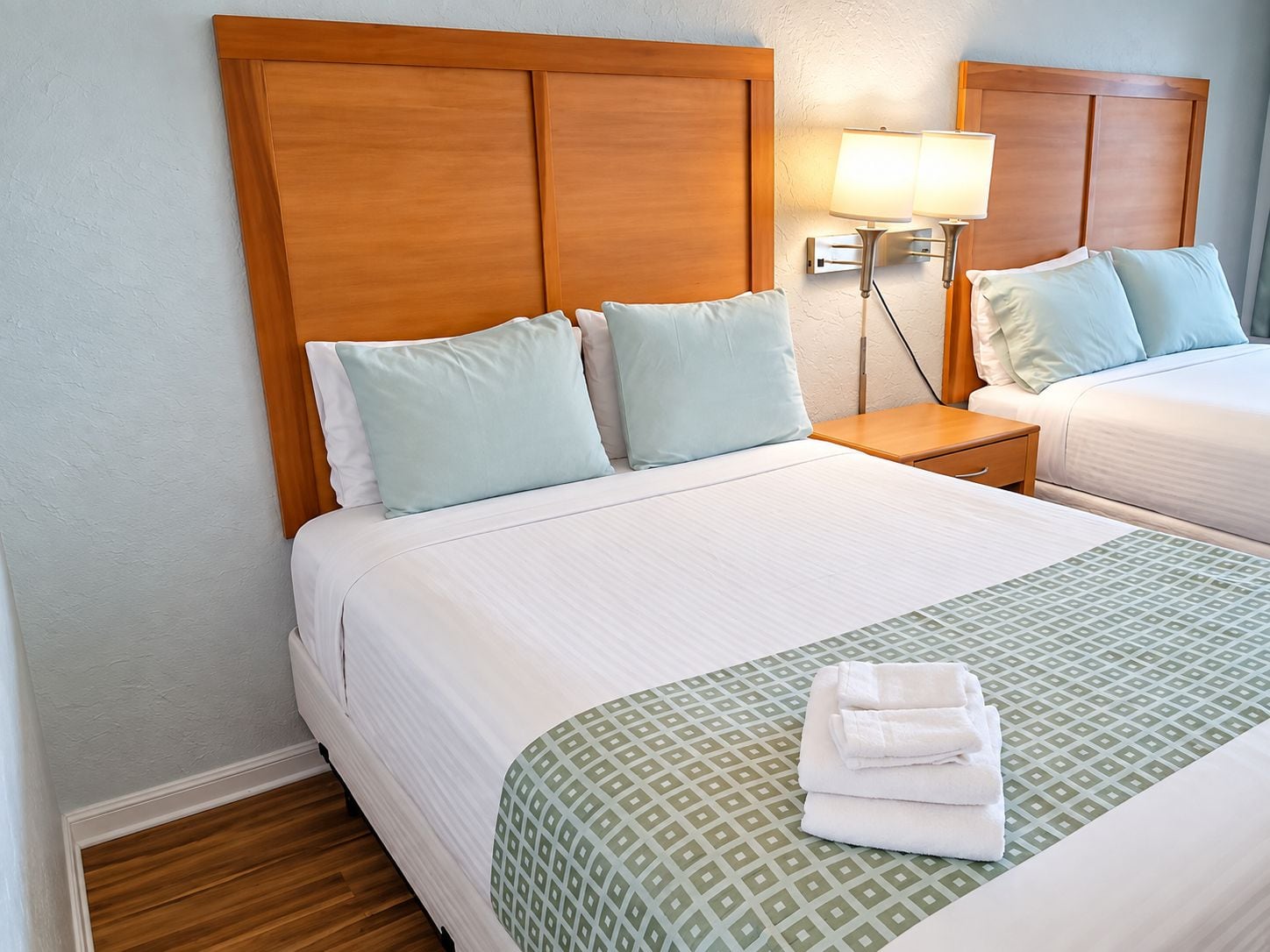Quarto de hotel com duas camas, cabeceira de madeira, mesa de cabeceira com abajur e colcha estampada.