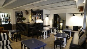 Uma área de bar com balcão listrado em preto e branco, prateleiras com garrafas e um espaço de estar com mesas e cadeiras.