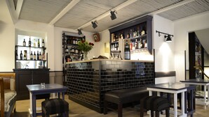 Uma área de bar com balcão revestido de azulejos pretos, várias prateleiras de garrafas e um espaço de estar com mesas e cadeiras.