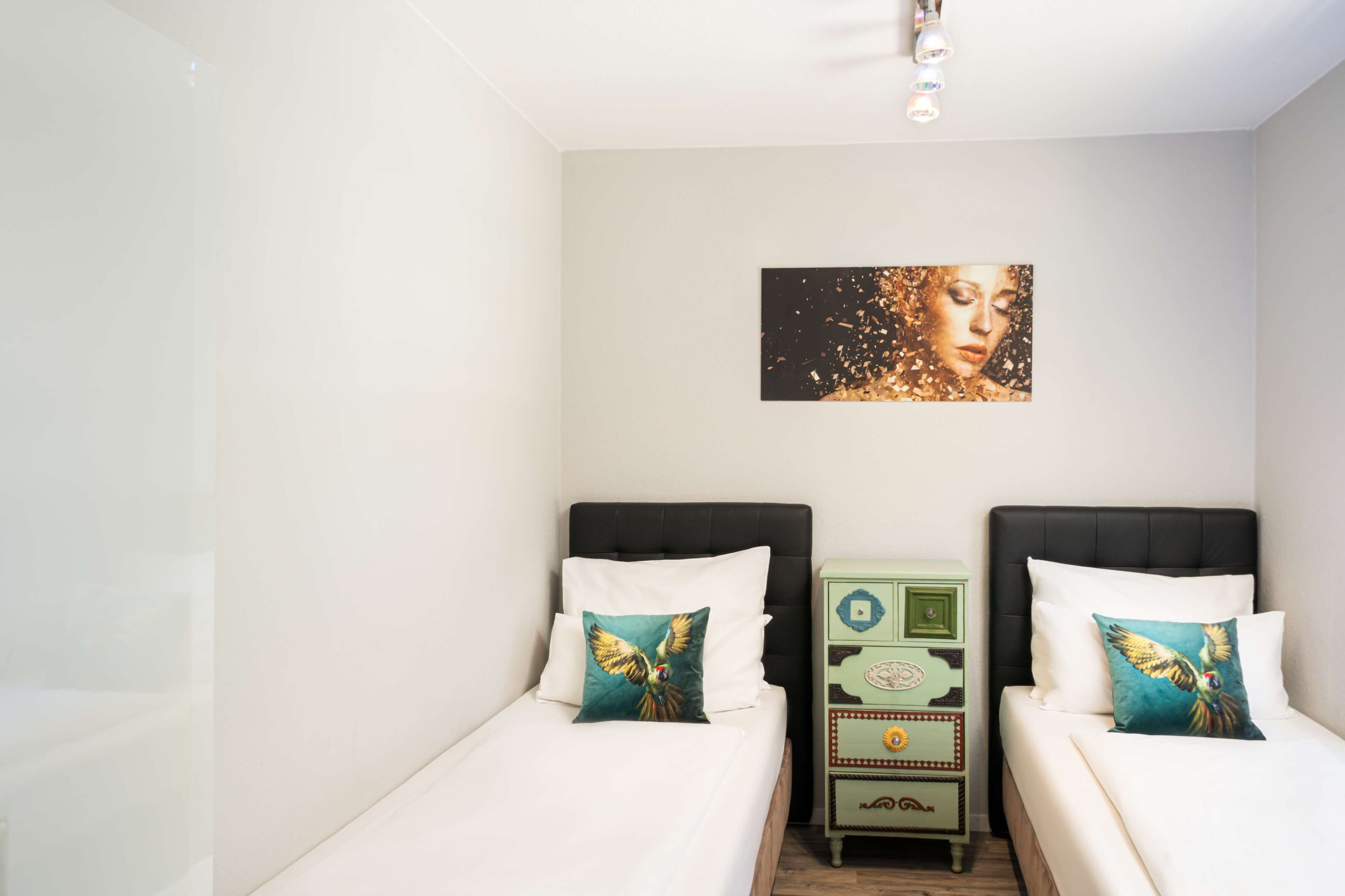 Quarto com duas camas de solteiro, cada uma com colchão e almofadas brancas. Entre as camas, um aparador verde com padrões decorativos.
